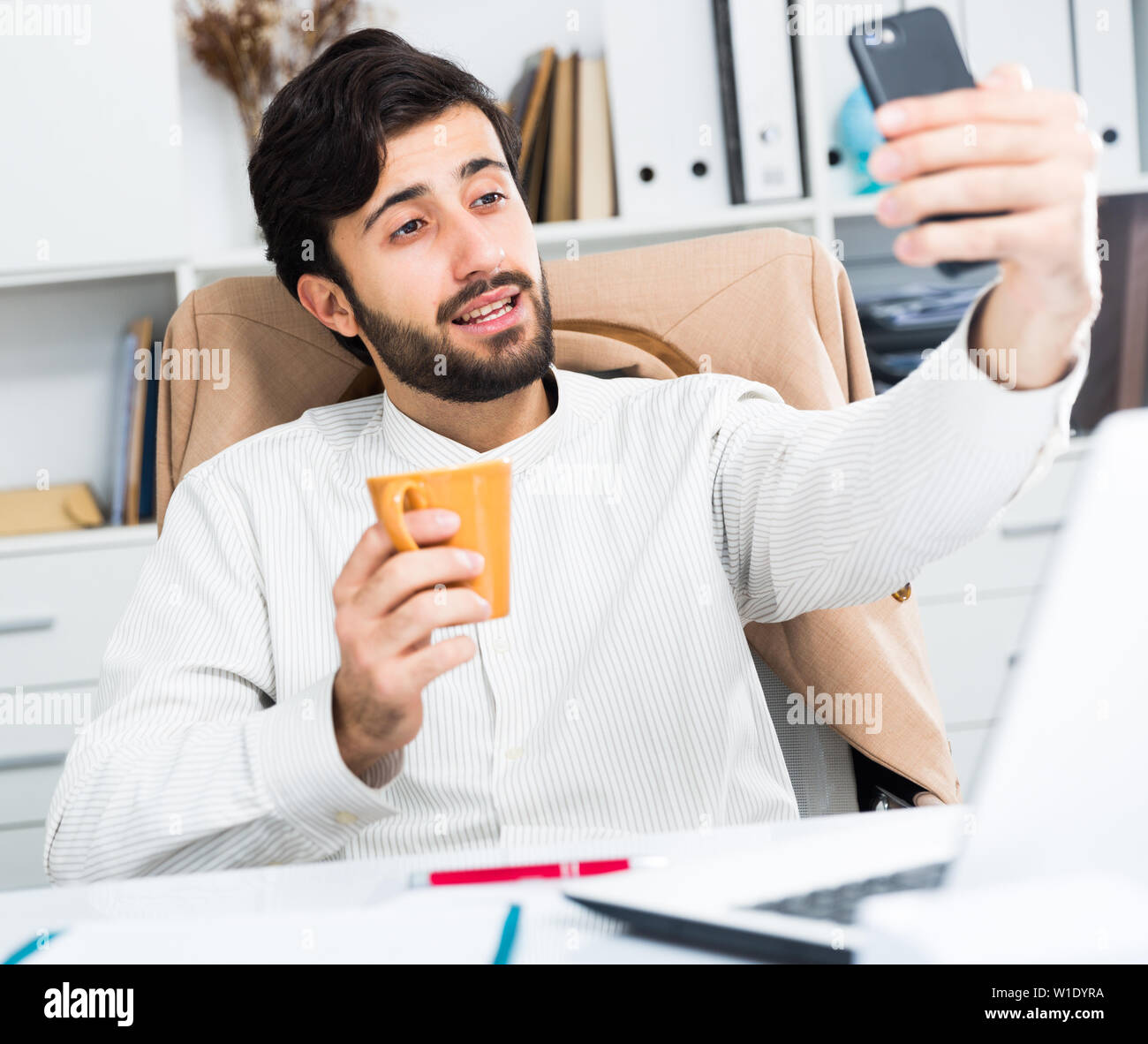 Cheerful young office worker taking selfie on his smartphone at desk in ...