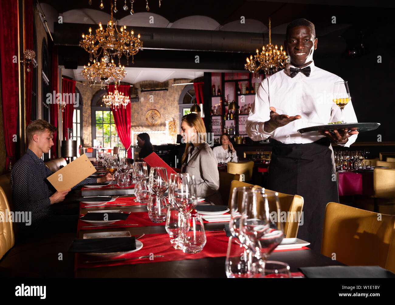 Polite waiter holding tray in restaurant with customers behind him ...