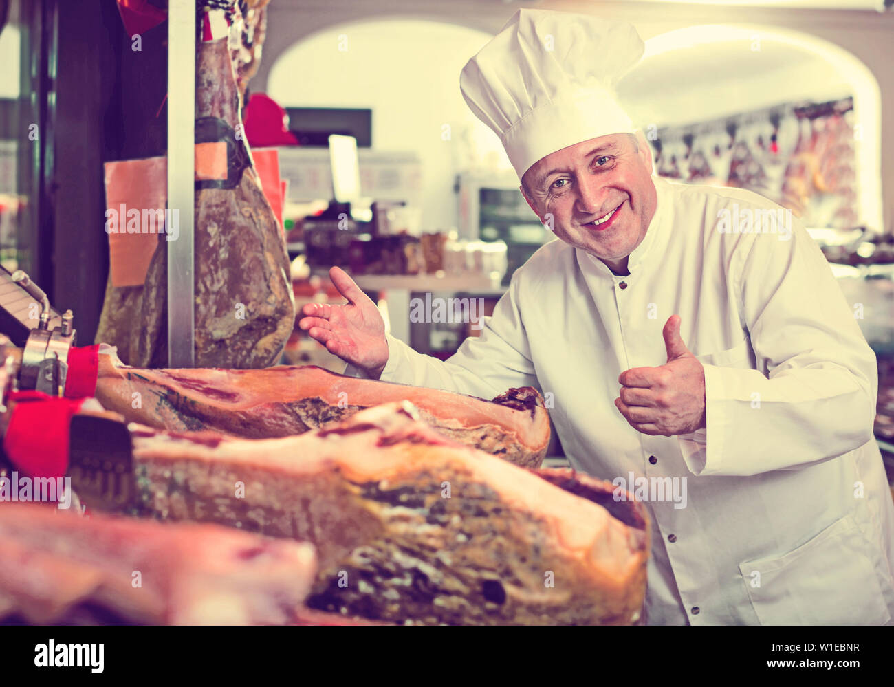 Portrait of smiling positive mature shop man offering Spanish jamon in ...