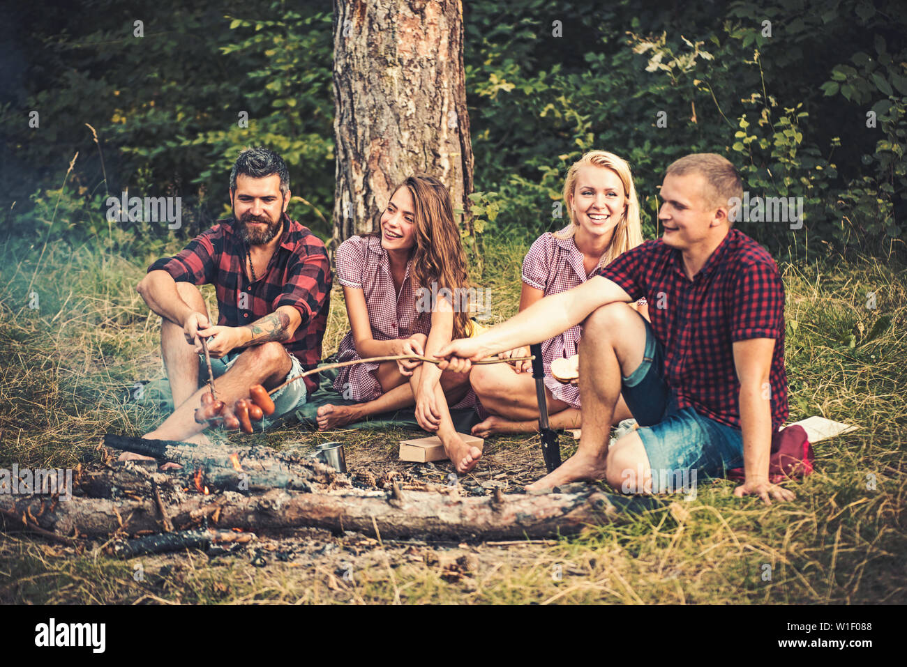 Two couples camping in woods. Guys frying sausages over fire. Smiling ...