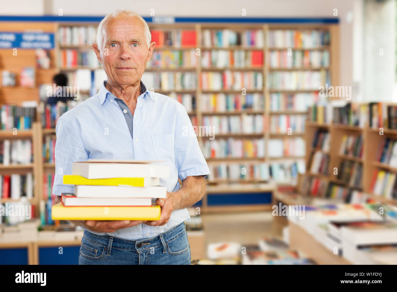 Elderly man holding stack of books near bookshelves in library interior ...