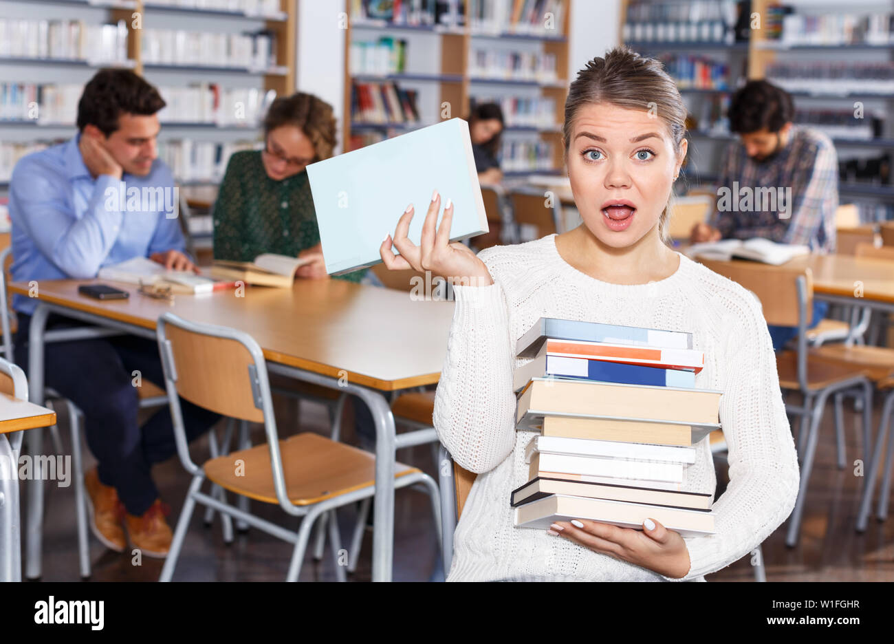 Surprised girl sitting with pile of books in public library on ...