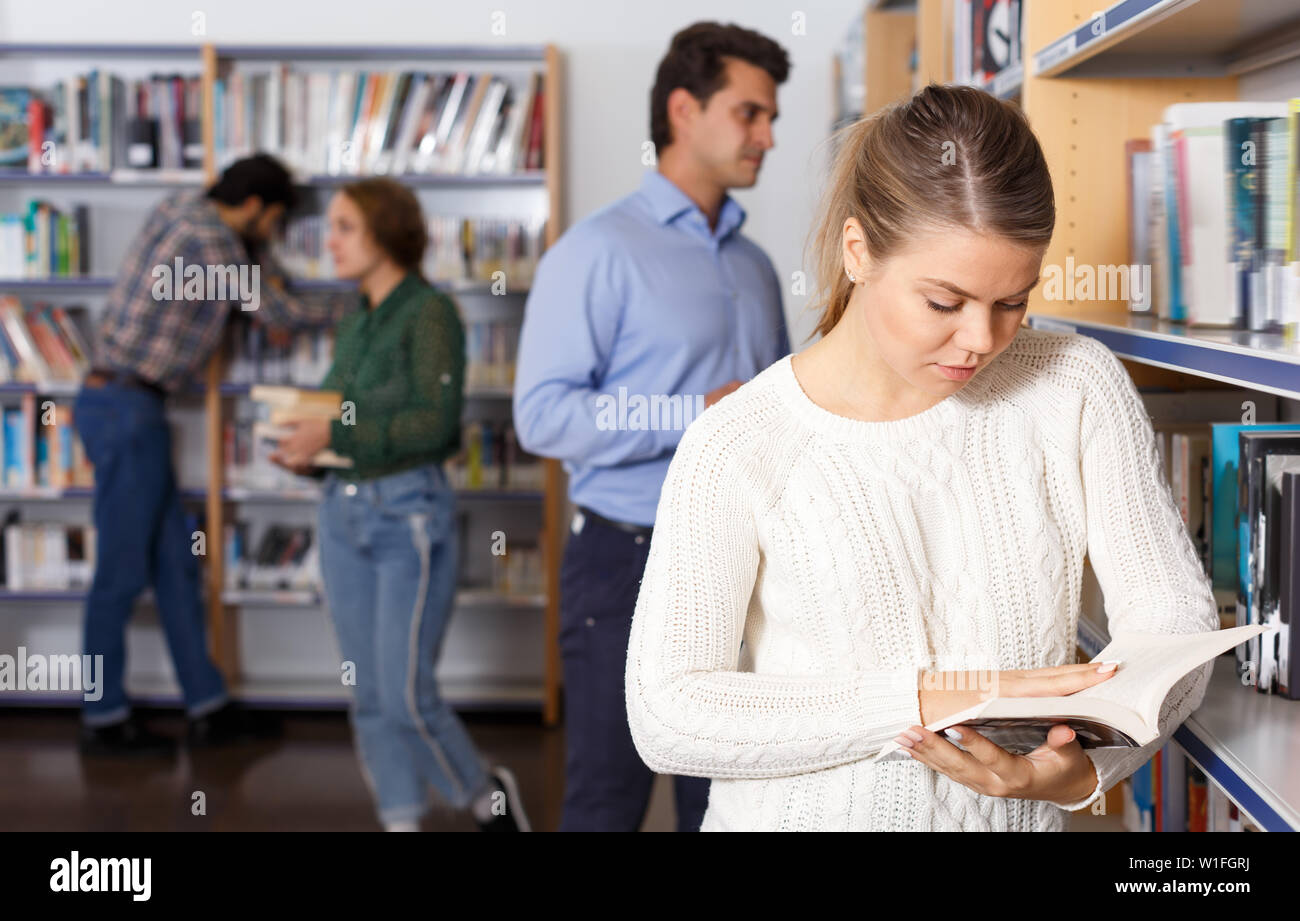 Adult attractive girl reading book while standing near bookshelves in ...