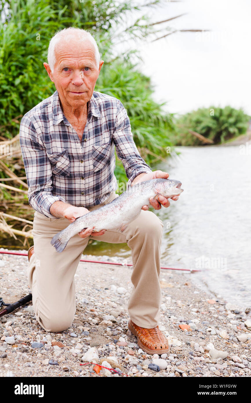Senior male fisher examining catch fish while angling at lakeside on ...