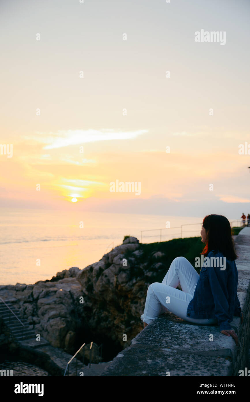young pretty woman sitting at cliff looking on beautiful sunset. summer ...
