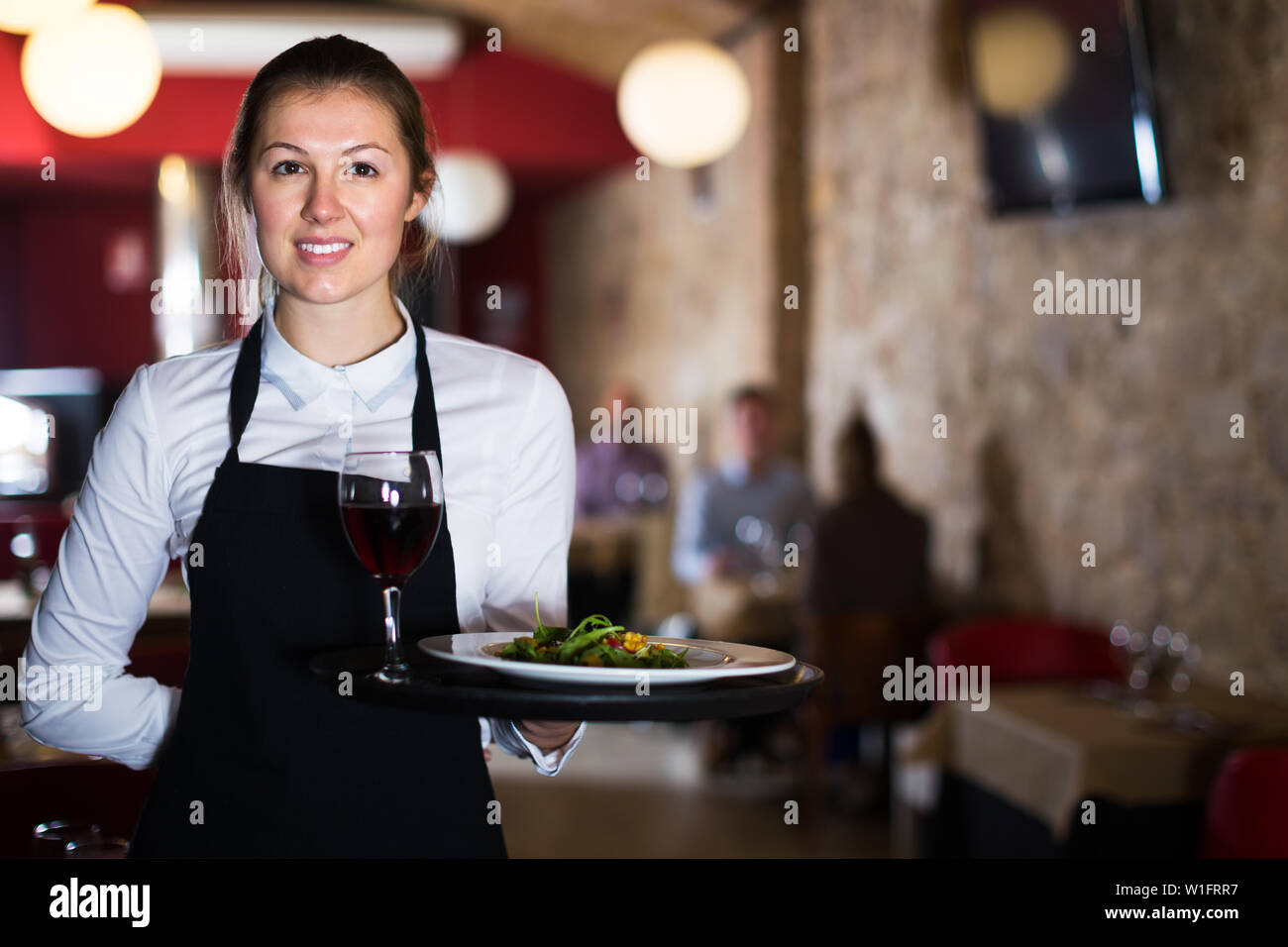 Portrait of smiling waitress wearing apron holding tray with dishes in ...