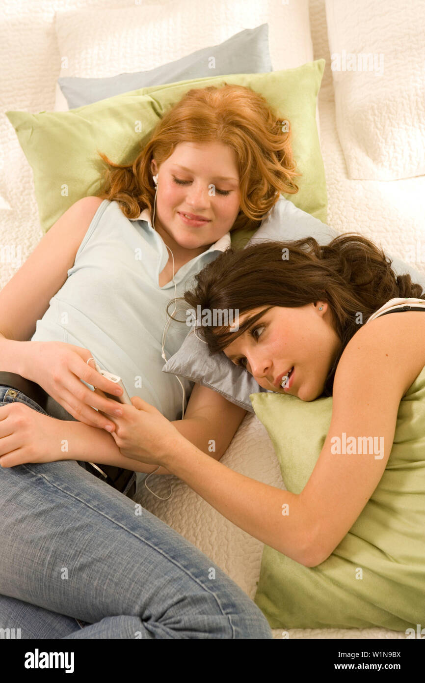 Two teenage girls (14-16) snuggling together on bed, listening to ...