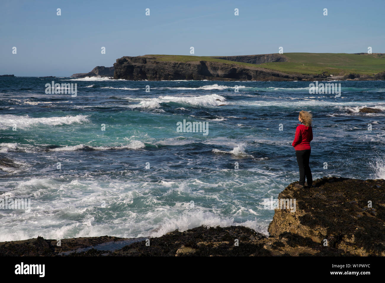 Waves of the Atlantic Ocean crash against rocks where a woman in a red ...