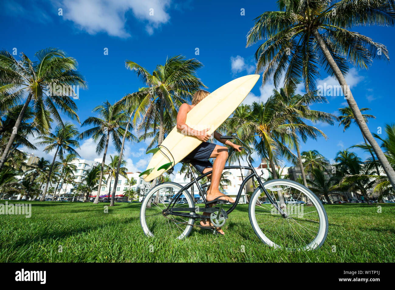Surfer with blond hair carrying his surfboard on a beach cruiser bike ...