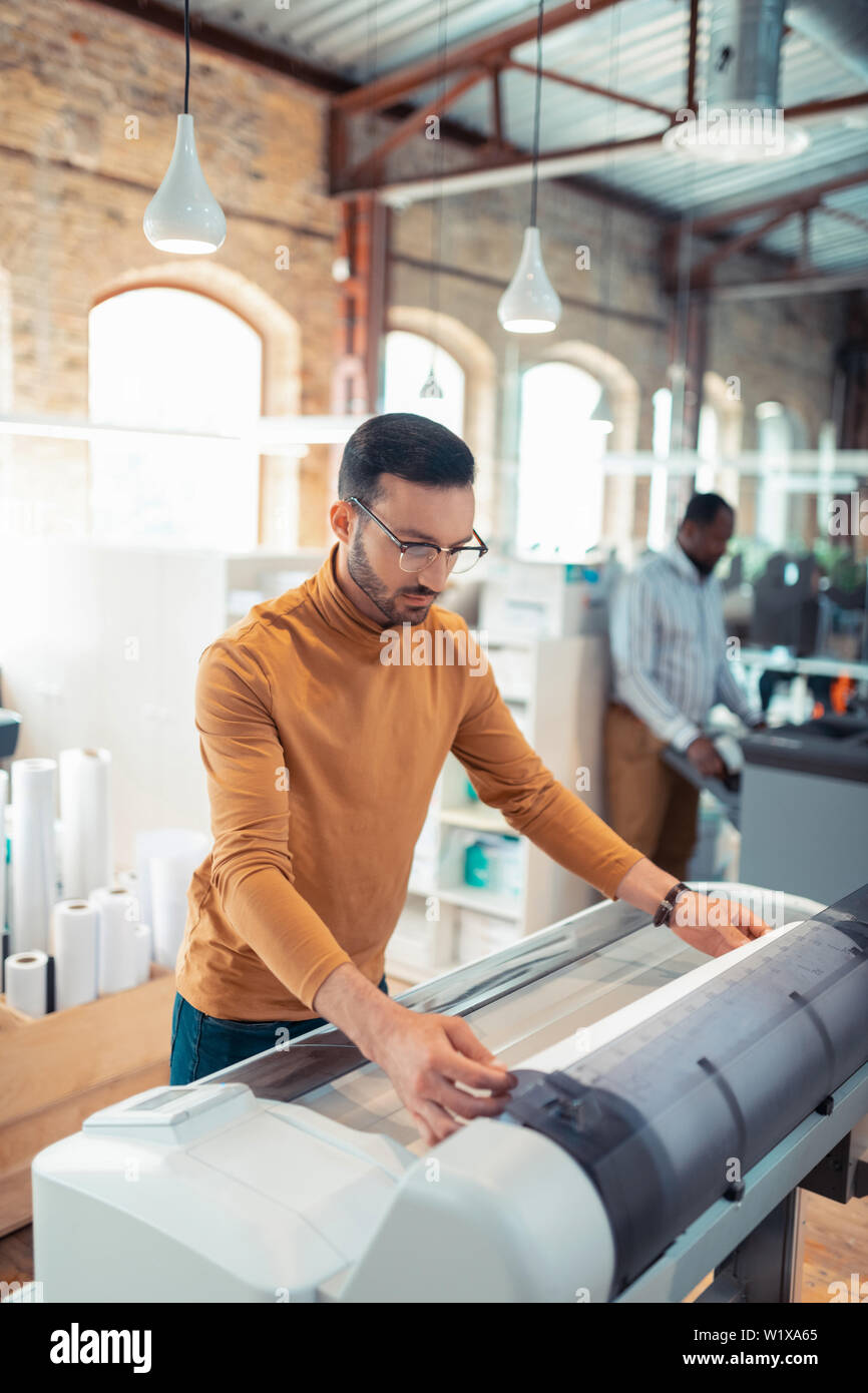 Using big printer. Bearded dark-haired man wearing glasses using big ...