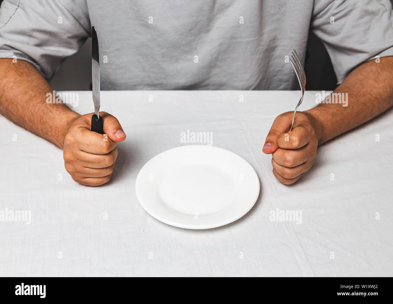 man sitting behind a table with fork and knife in hands and empty plate ...