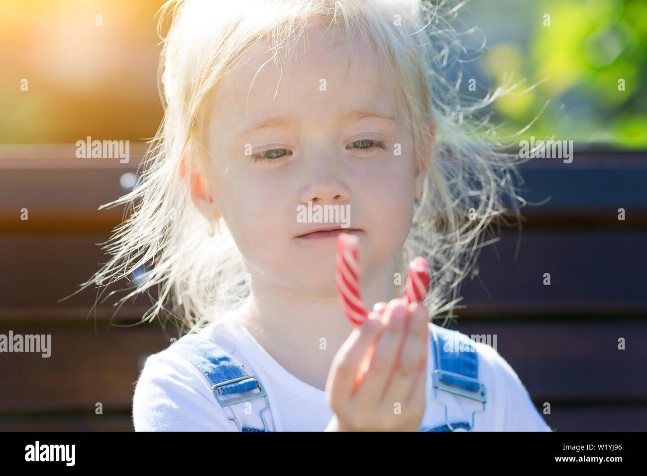 A little girl sucks a big sweet candy while sitting on a bench city ...