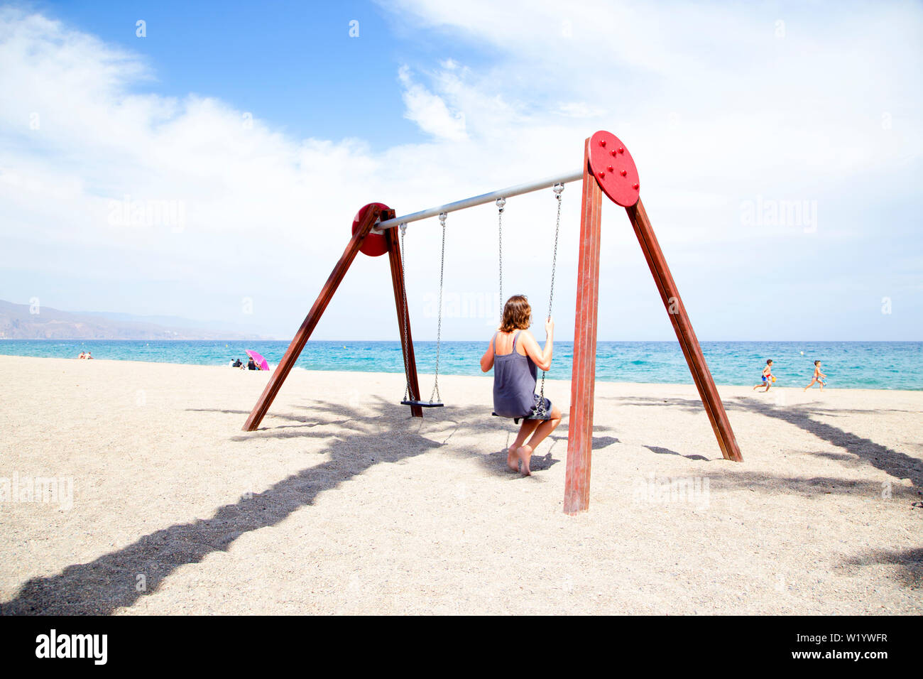 middle aged woman sitting on a swing. On the beach with the sun on her ...