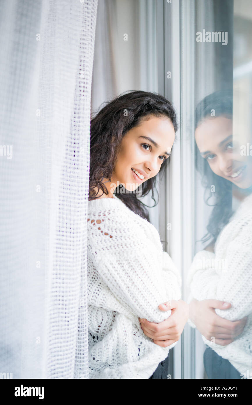 Beautiful young woman looking through window while sitting at ...