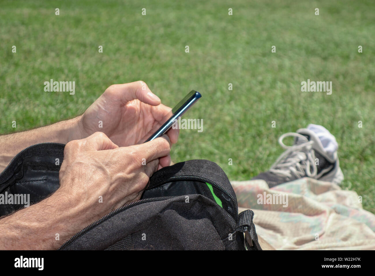 Man using his mobile phone on a picnic blanket at a public lawn park ...