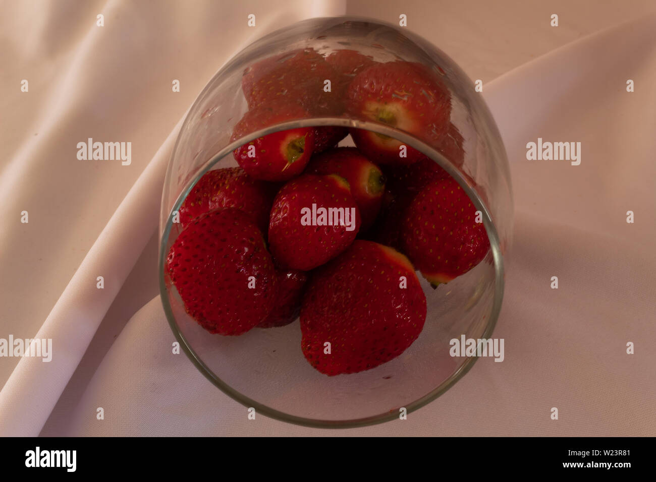 Many red strawberries inside a glass cup, taken with a white fabric ...