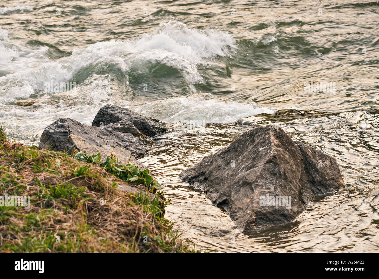 Water flowing near rock at shore, forming small wave in river, lit by ...
