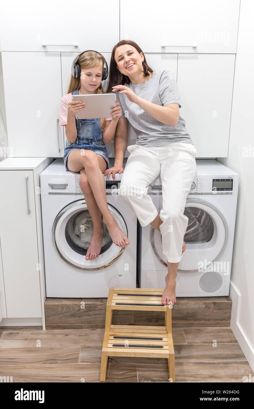 Young Mother And Her Daughter Are Playing With Tablet In Laundry Room