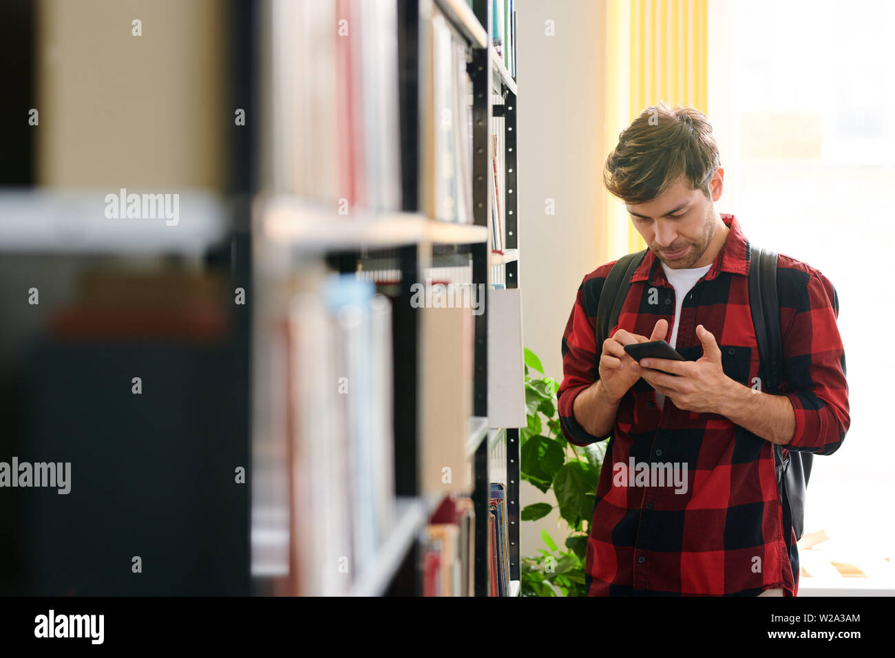 Young man scrolling in smartphone by shelf in library Stock Photo - Alamy