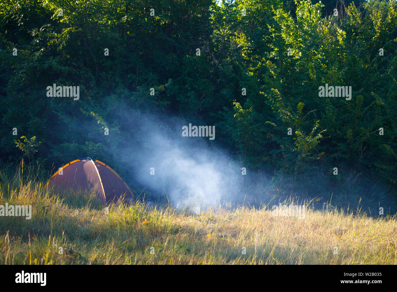 Tourist tent and smoking bonfire in a field in front of the forest at ...