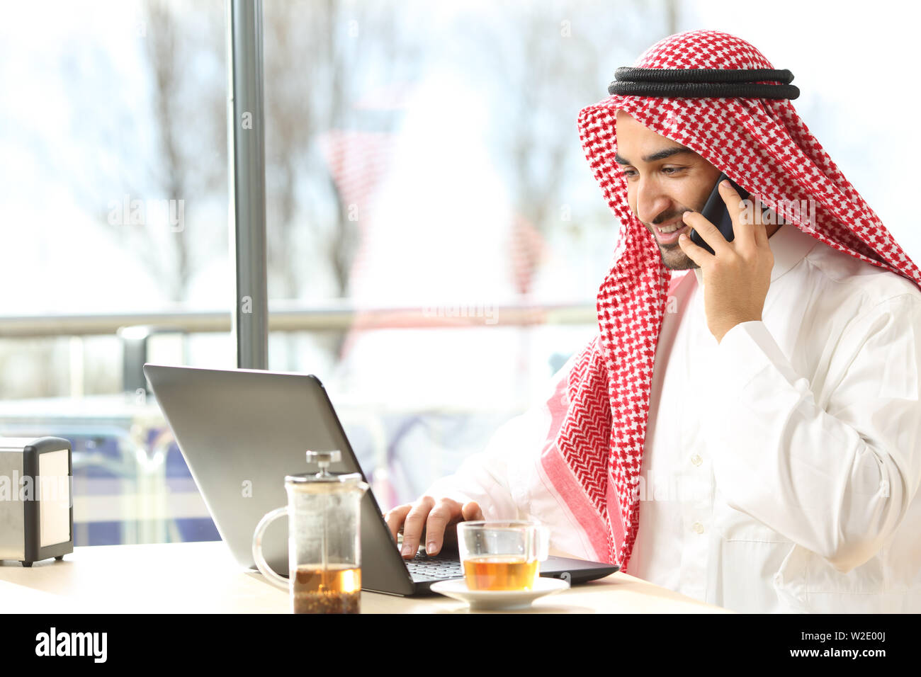 Happy arab man using a laptop and talking on smart phone sitting in a ...