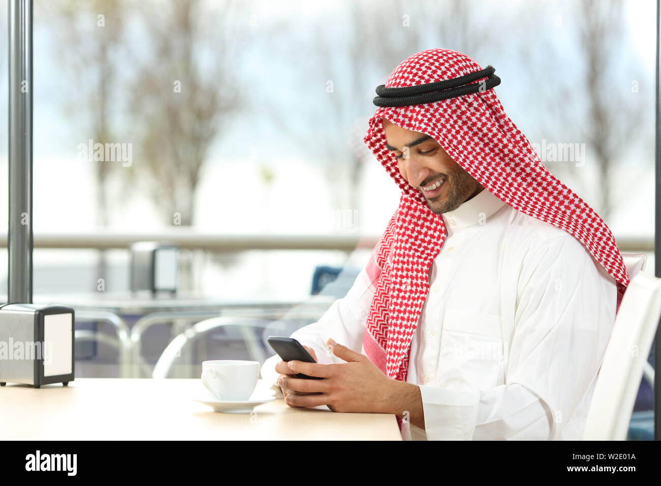 Happy arab man using a smart phone sitting in a coffee shop interior ...