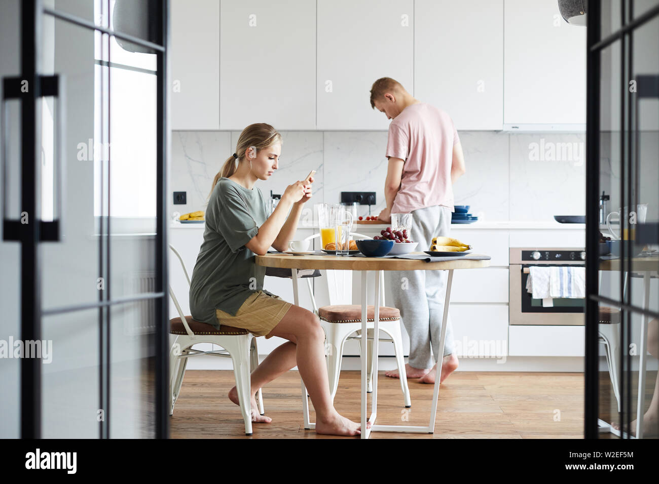 Young wife texting in smartphone by brunch while her husband cooking in ...