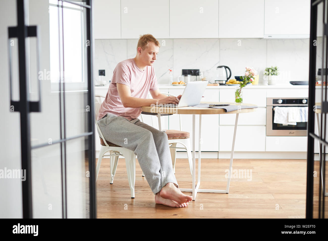 Young casual man sitting by table in the kitchen in front of laptop ...