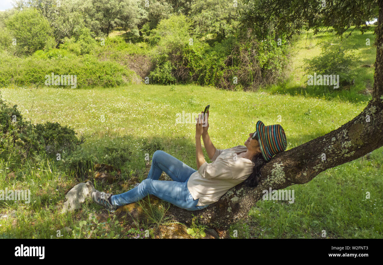 Brunette woman sitting on a tree in a green grass field, looking at her ...