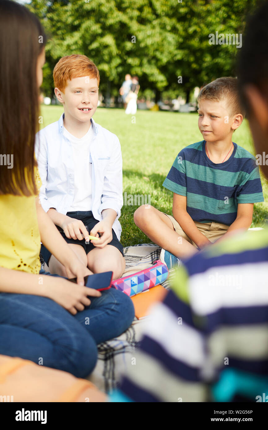 Four schoolchildren sitting on grass in park after school, talking and ...