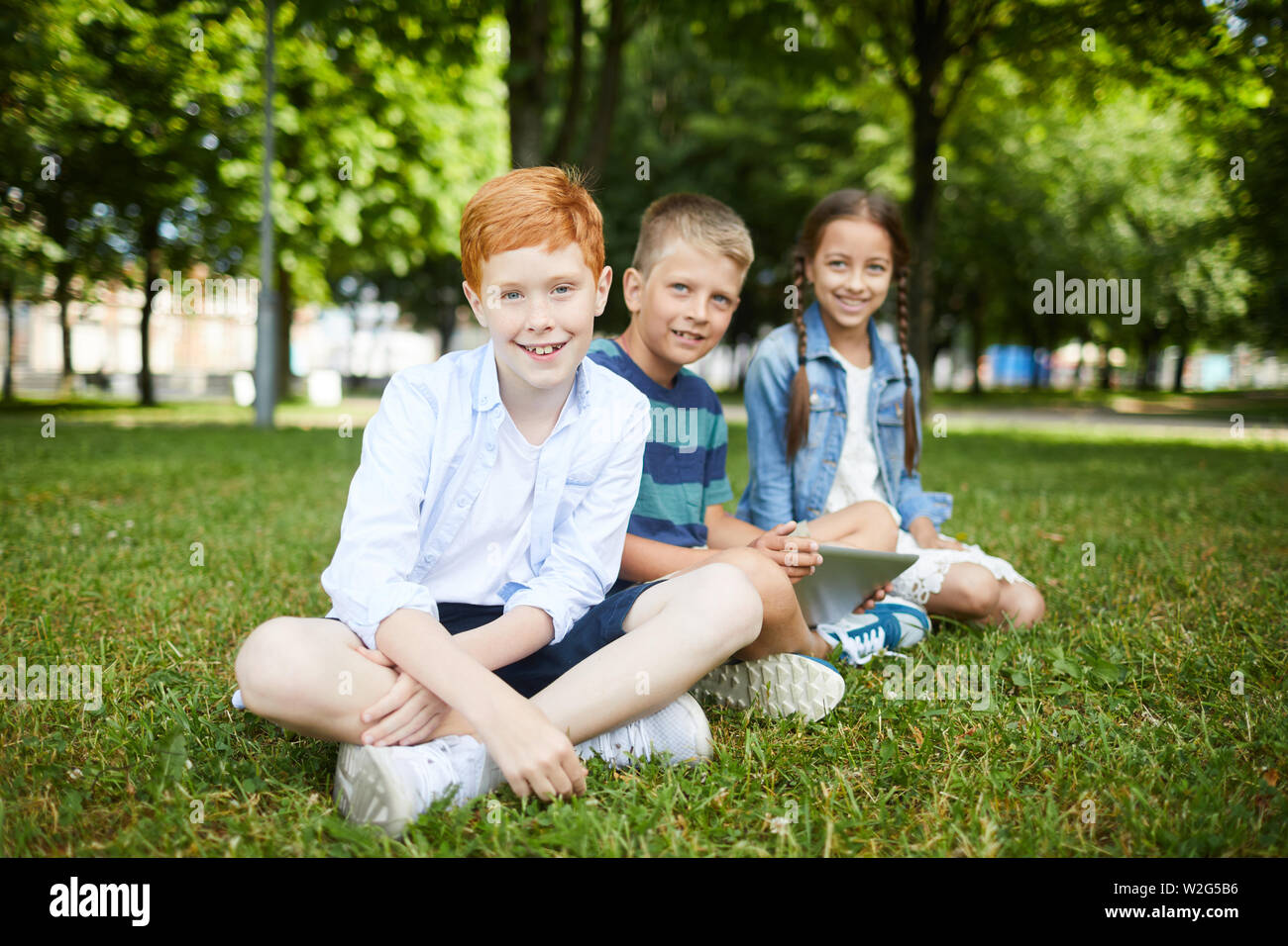 Three smiling schoolkids in park sitting on grass, having fun together ...