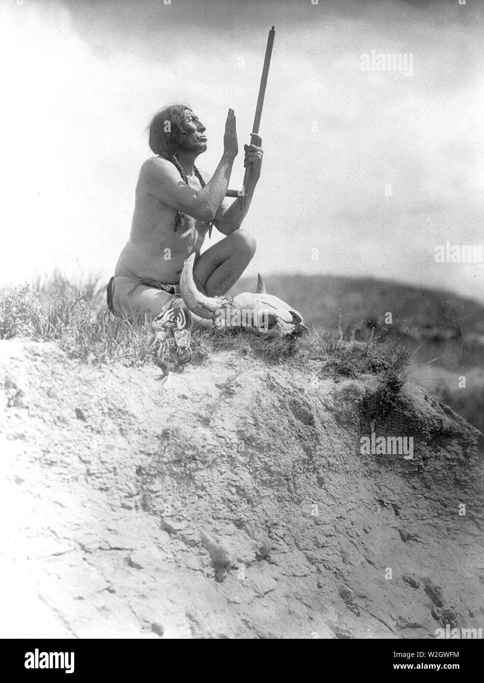 Slow Bull, squatting, wearing breechcloth, holding pipe with mouthpiece ...