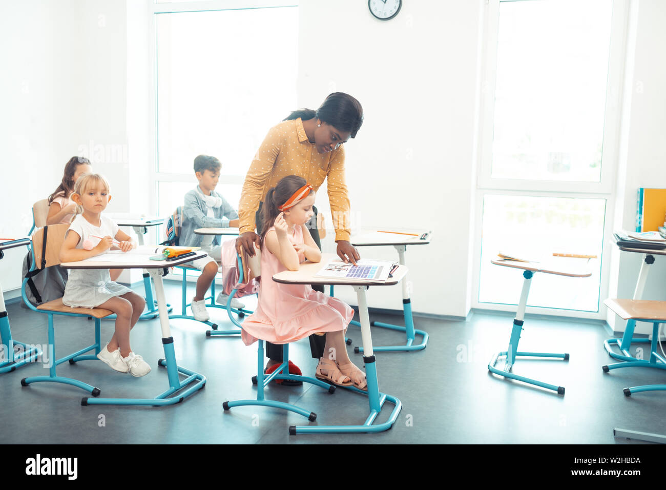 Helpful dark-skinned teacher helping children with task Stock Photo - Alamy
