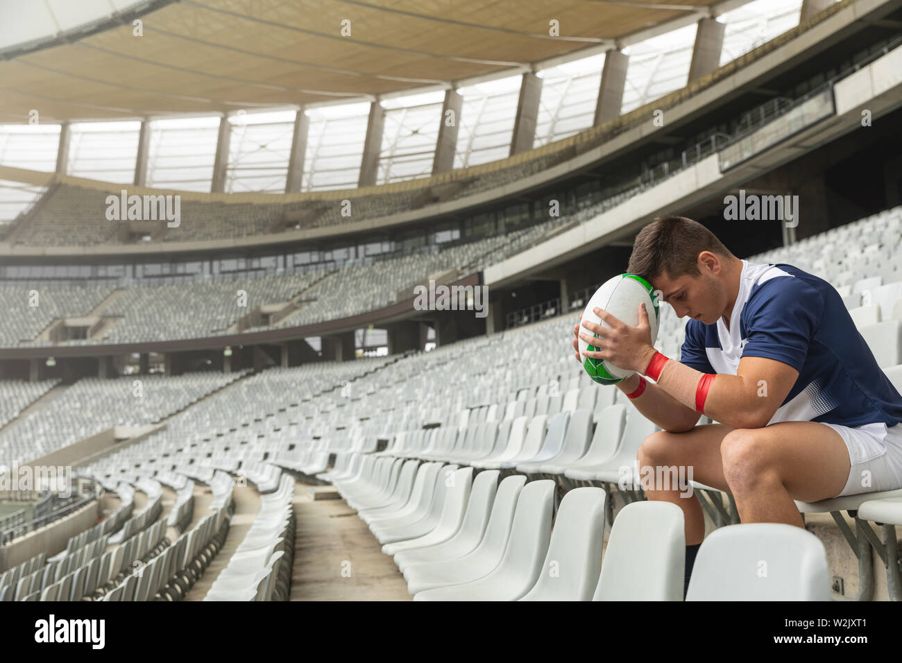 Upset Caucasian Male rugby player sitting with rugby ball in stadium ...
