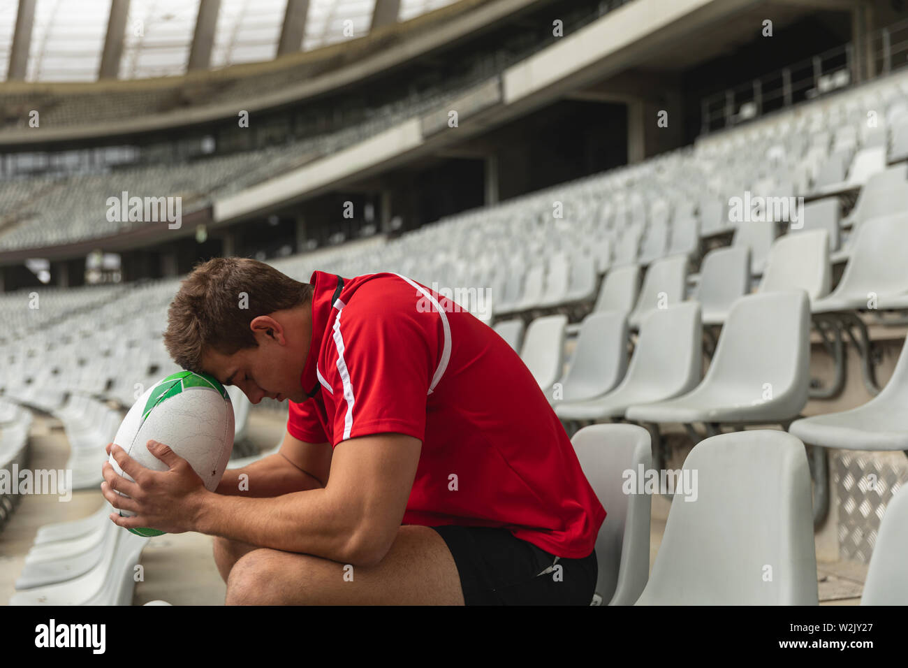 Upset Caucasian male rugby player sitting with rugby ball in stadium ...