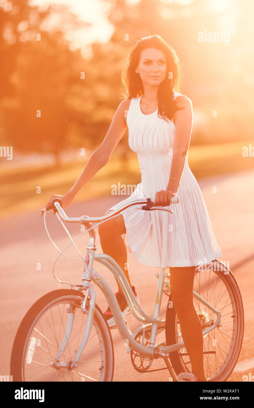 Beautiful brunette girl in white dress on bicycle at sunset in park ...