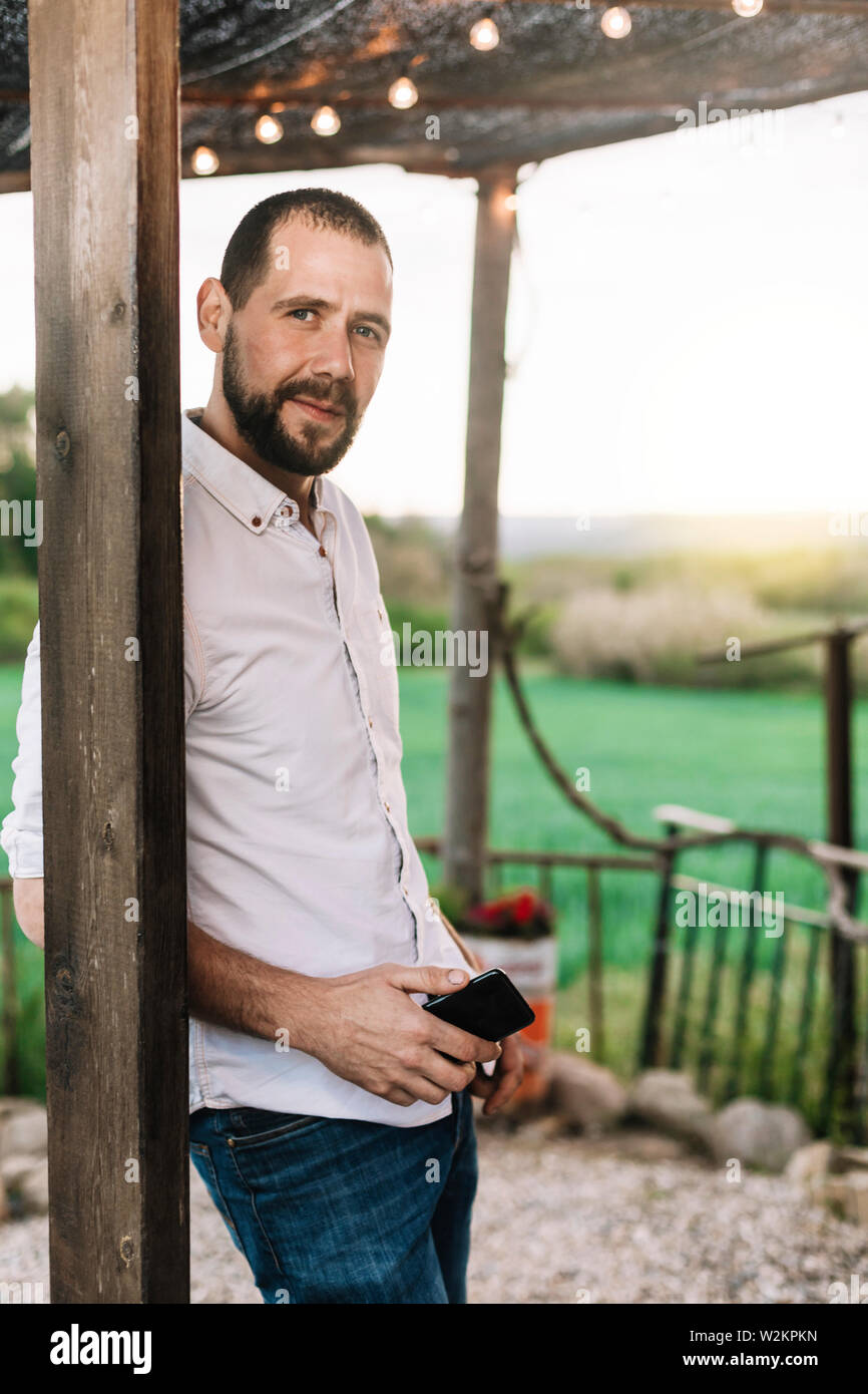 Bearded man standing at countryside home while using a mobile phone ...