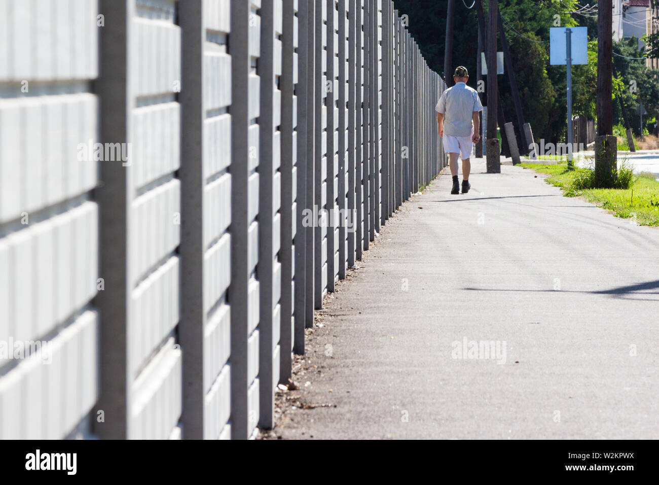 Elderly man walking on asphalt pavement beside concrete fence in the ...