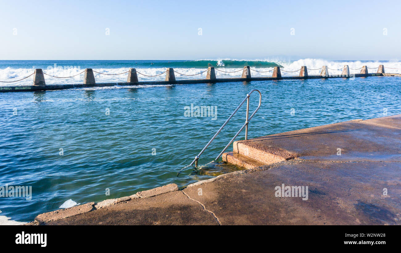 Beach tidal swimming pool with surfers surfing ocean waves breaking ...