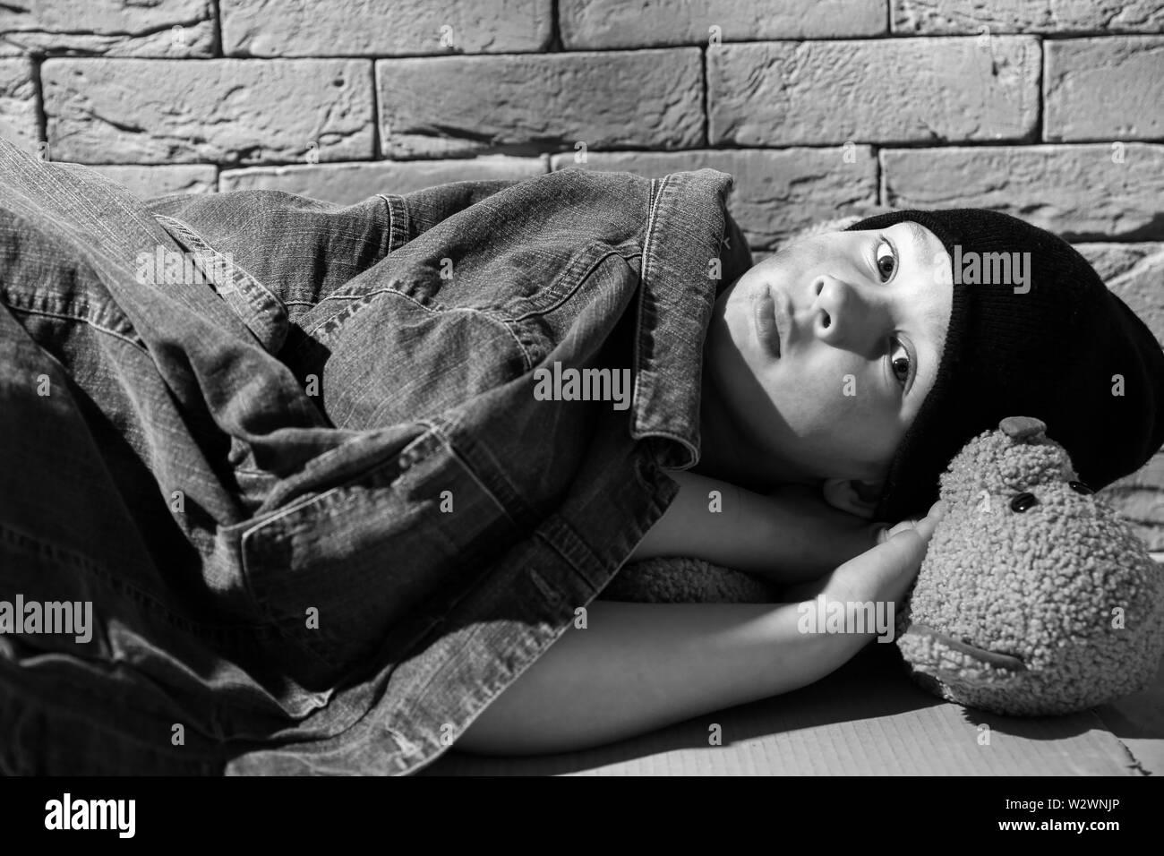 Homeless little boy with teddy bear lying on floor near brick wall ...