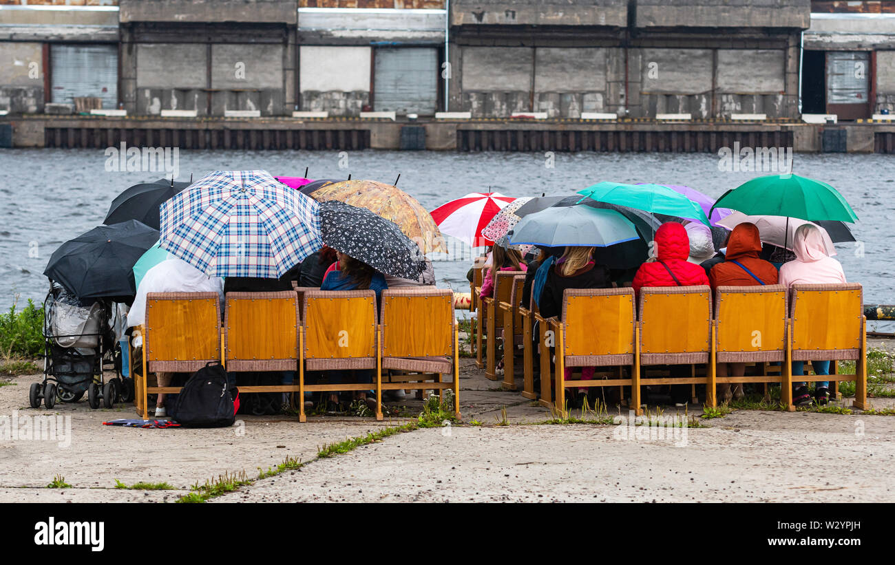 The crowd of people with colorful umbrellas sit on the benches at the ...