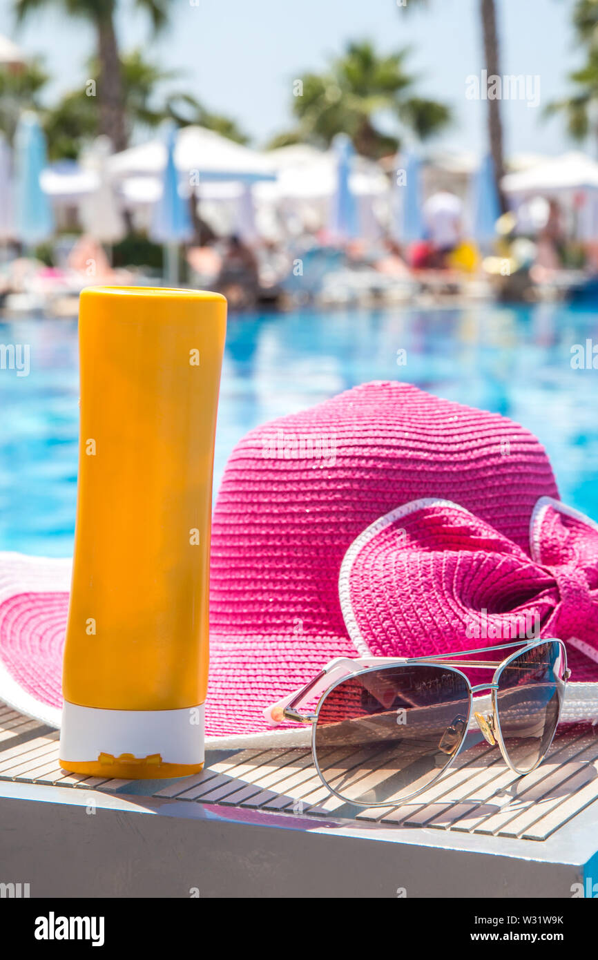 Bottle of sunscreen, hat and sunglasses next to swimming pool in hotel ...
