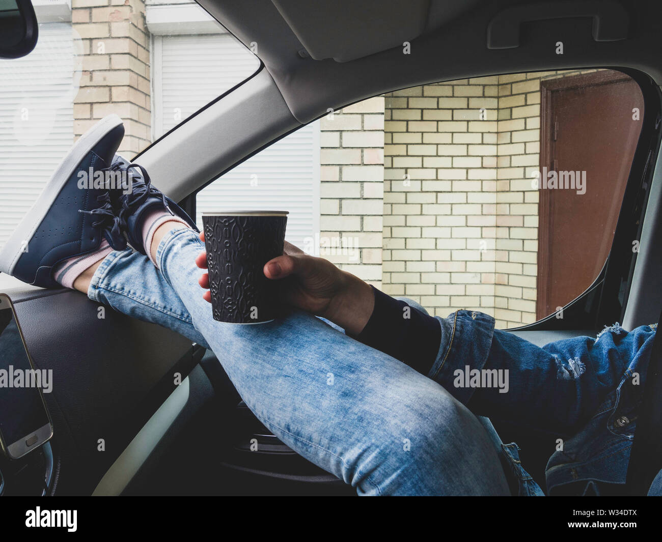 Young drinking coffee take away with feet in boots on car dashboard ...