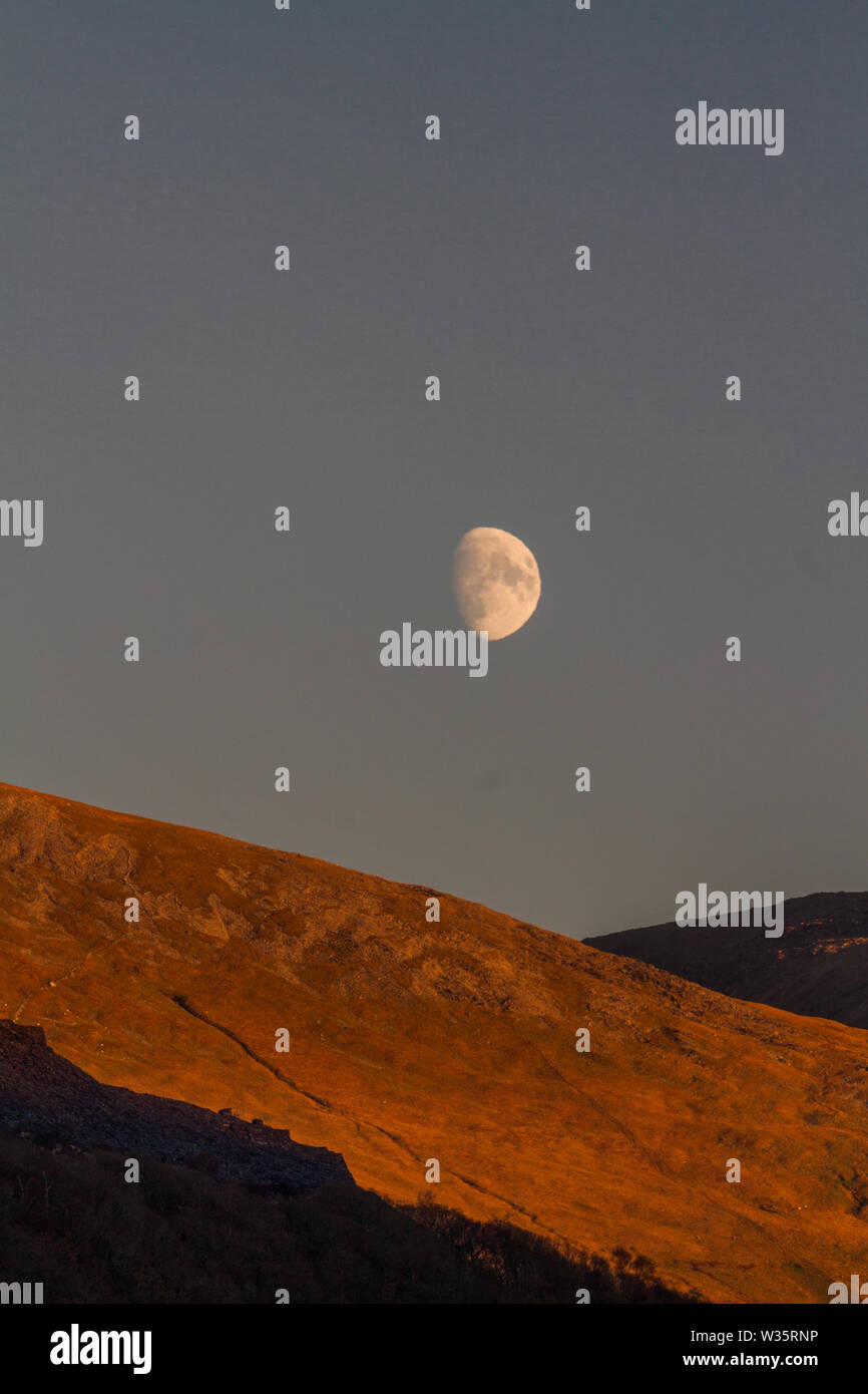 Nearly full moon rising over hill that is catching last light of day ...