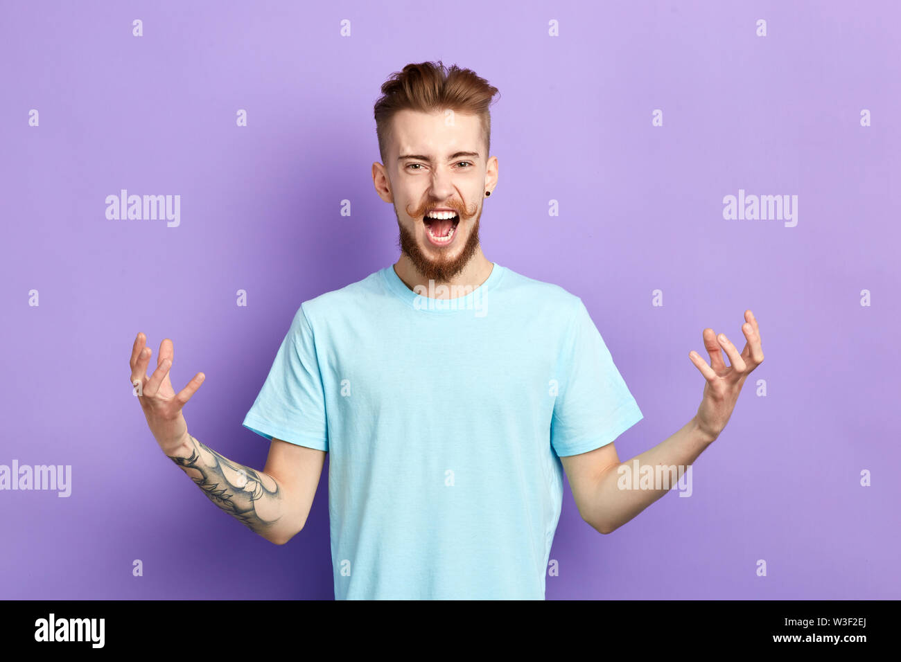 angry young man with moustache in blue shirt standing with raised arms ...