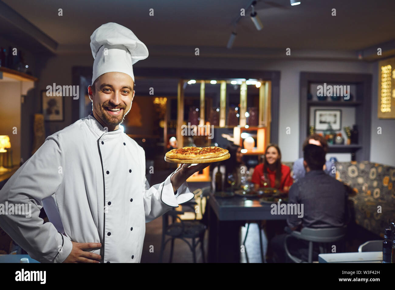 A male cook is smiling against the background of people at a table in a ...
