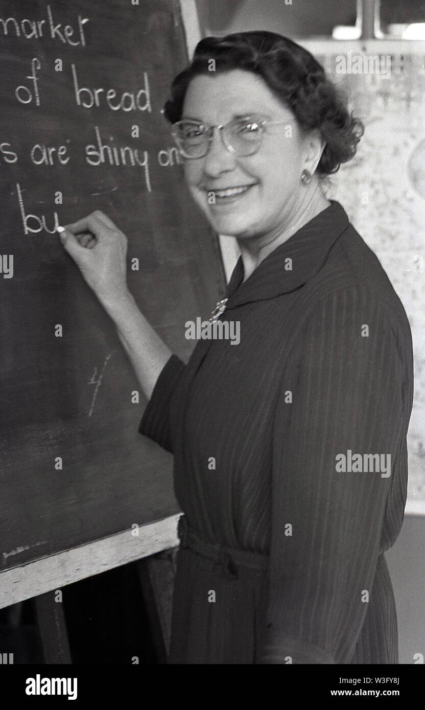 1950s, historica, a smiling lady teacher writing with chalk at her ...