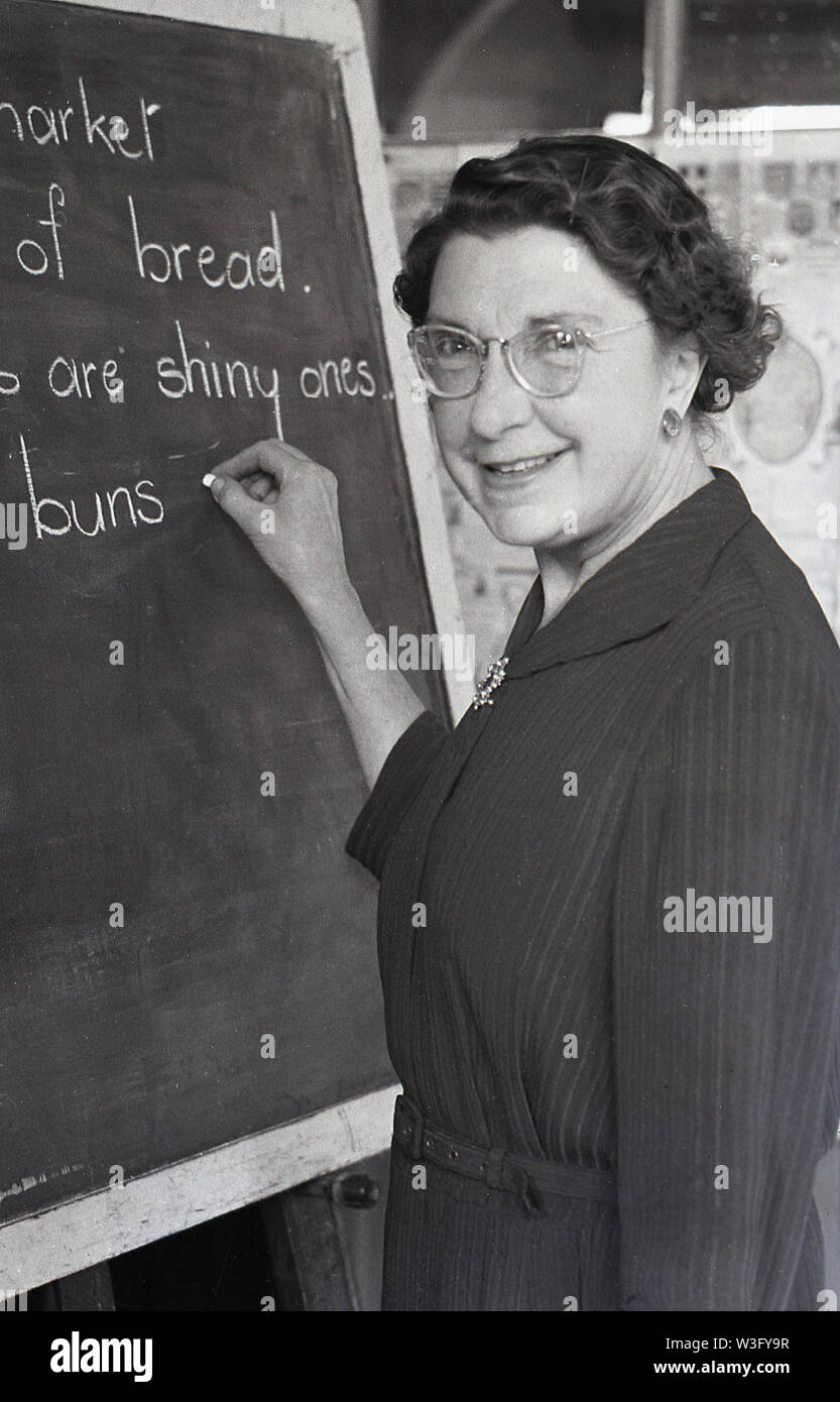 1950s, historica, a smiling lady teacher writing with chalk at her ...