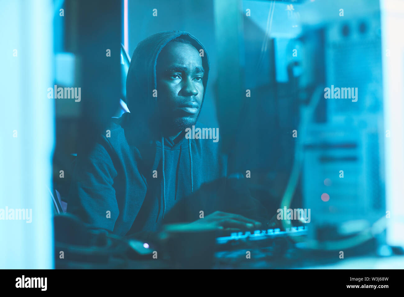 Serious tired black man in hoodie sitting in dark blue room and typing ...