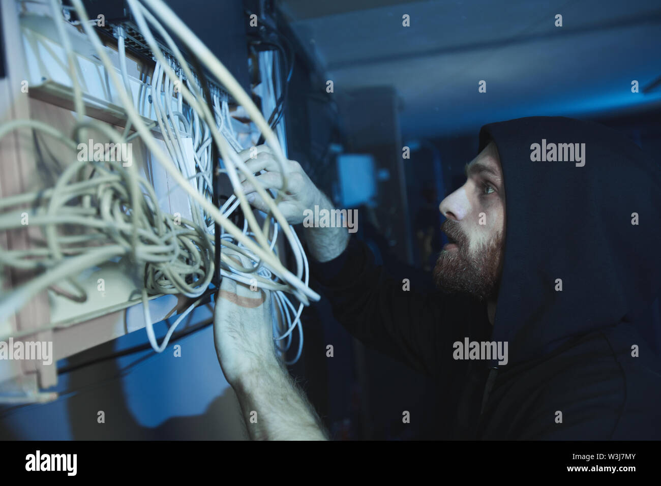 Concentrated young bearded hacker in hoodie checking cables of network ...
